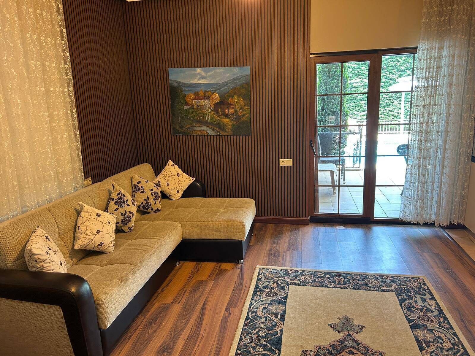 Sala de estar com sofá, almofadas estampadas, tapete, quadro e uma porta de vidro deslizante para uma área externa.