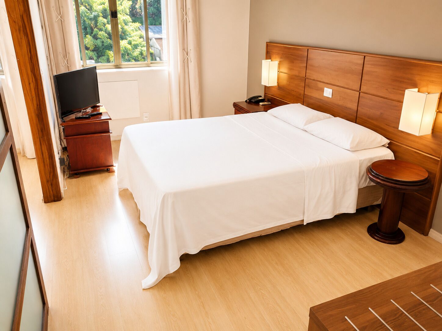 Quarto de hotel com cama, luminárias de cabeceira, mesa de cabeceira, televisão e janela com cortinas.