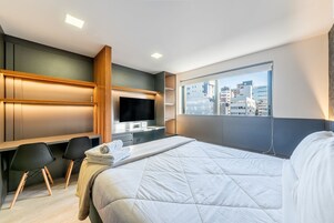 Quarto de hotel moderno com cama, escrivaninha, televisão e janela com vista para a cidade.