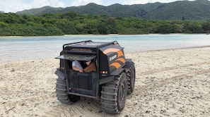 Um veículo numa praia arenosa, com um corpo de água e montanhas ao fundo.