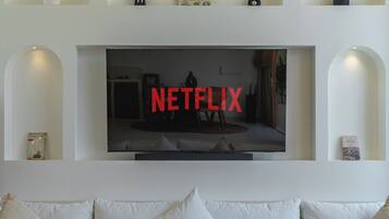 Uma sala de estar moderna com um sofá branco, mesas de apoio de vime e uma TV fixada na parede exibindo 'NETFLIX'.