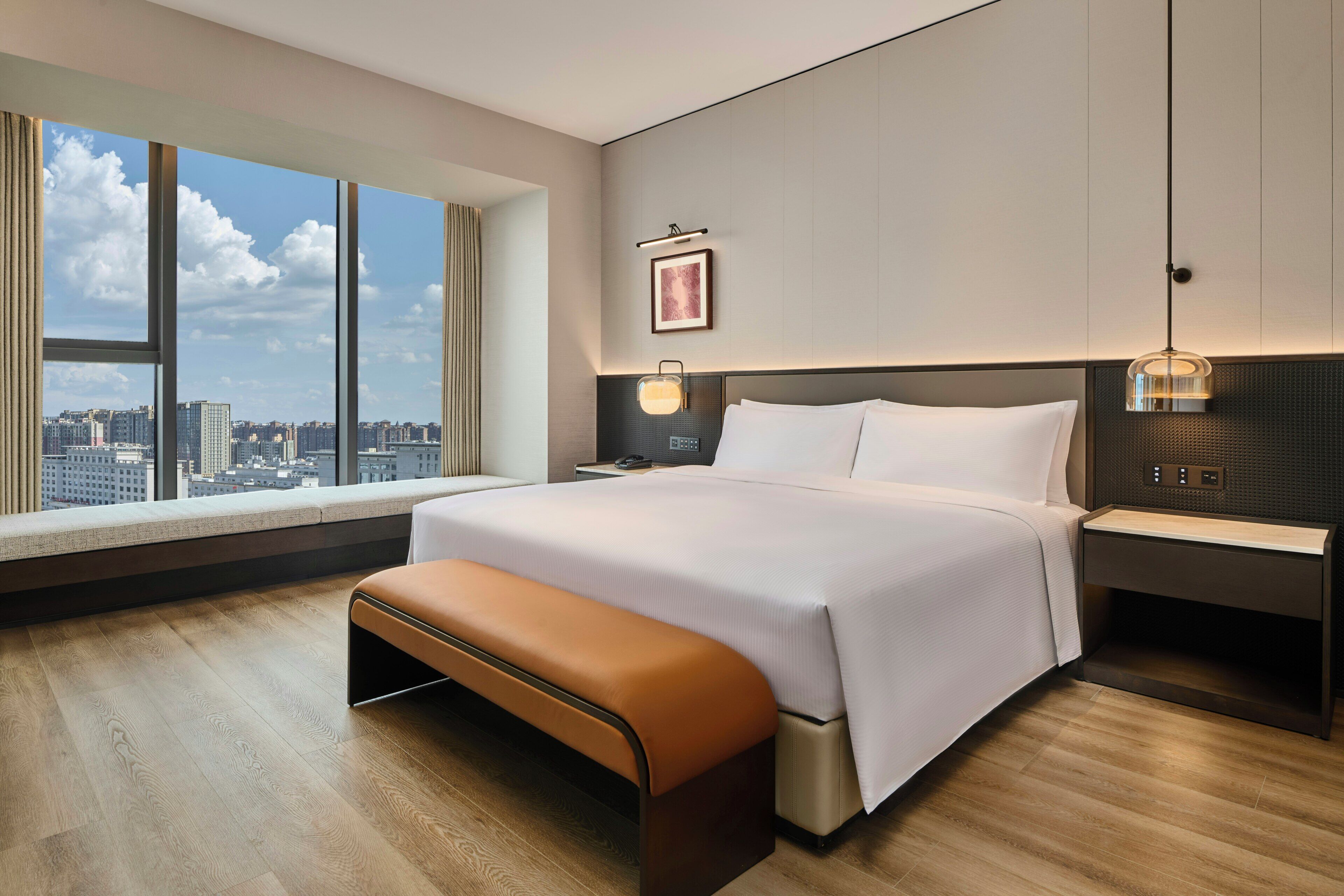 Quarto de hotel moderno com uma cama grande, um banquinho, uma mesa de cabeceira, uma luminária na parede e vista para a cidade.