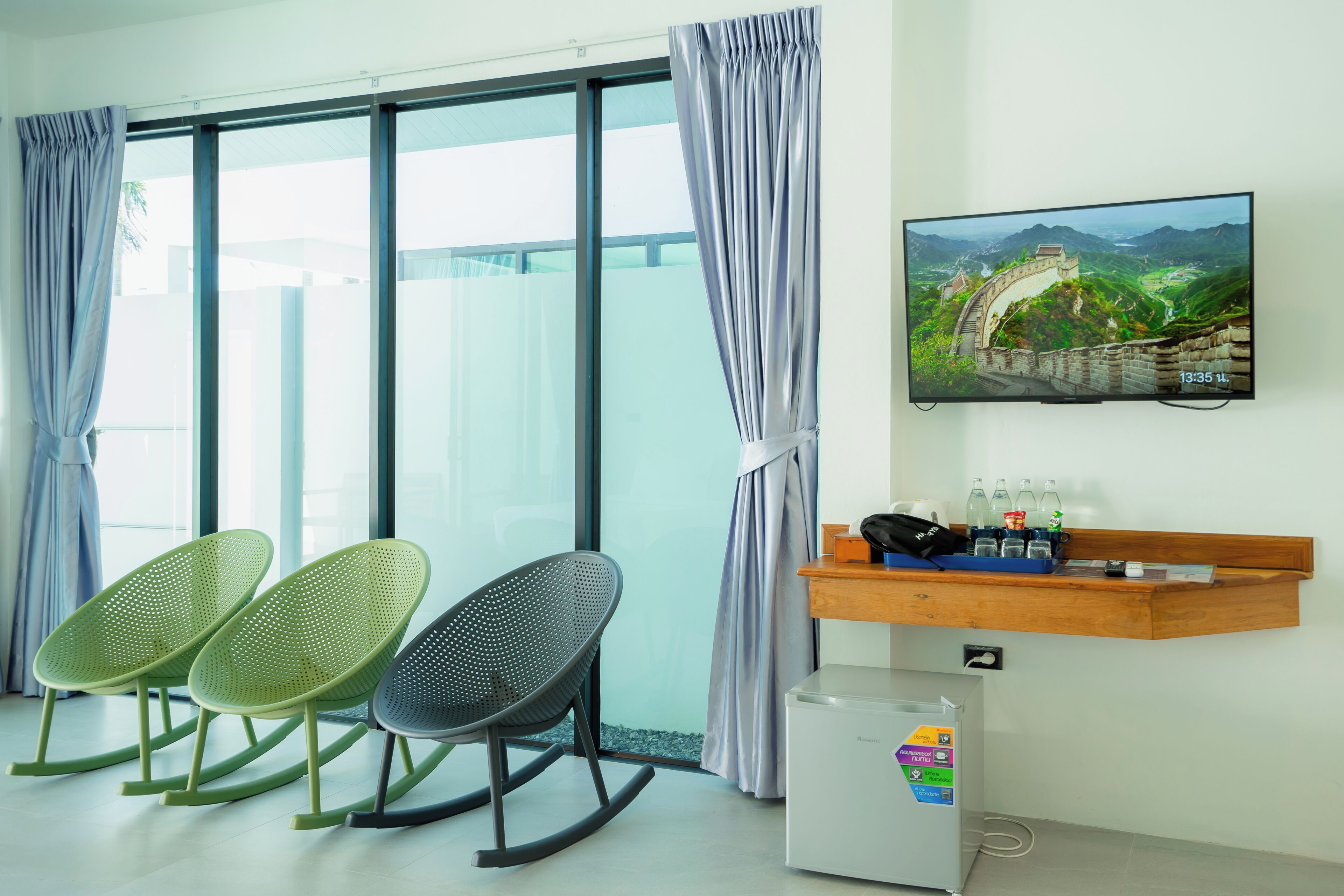Quarto com janelas amplas, televisão, frigorífico e duas poltronas de baloiço.