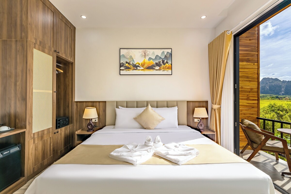 Quarto de hotel com uma cama grande, duas mesas de cabeceira com abajures, uma obra de arte na parede e uma varanda com vista para o verde.