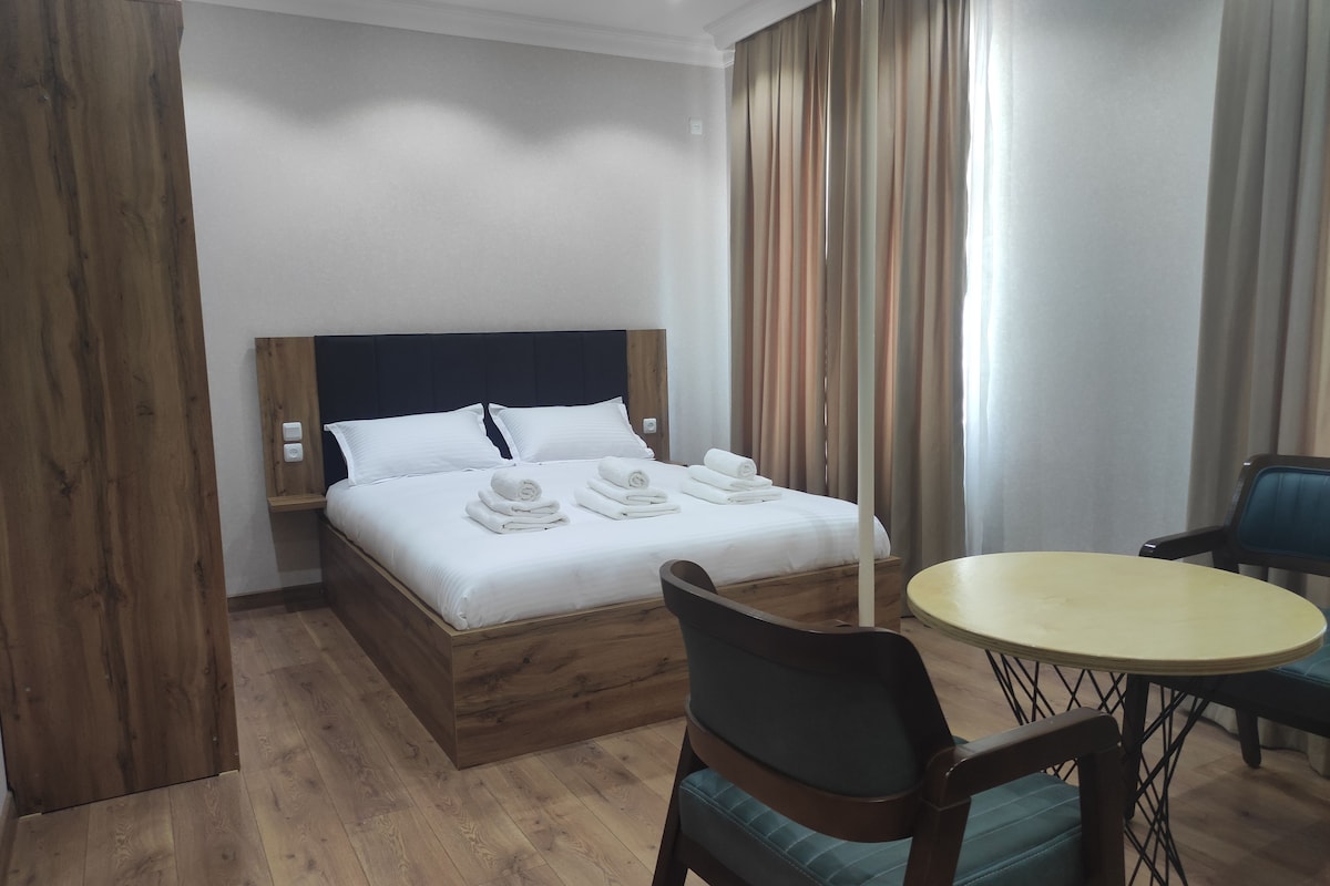 Quarto de hotel com cama, mesa de cabeceira, uma cadeira, uma mesa redonda e cortina.