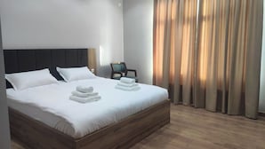 Cama bem arrumada com roupa de cama e toalhas brancas, cabeceira de madeira e uma cadeira em quarto com piso de madeira e cortinas.
