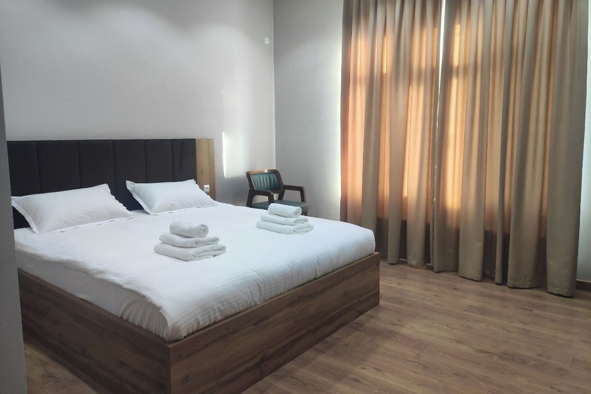 Cama bem arrumada com roupa de cama e toalhas brancas, cabeceira de madeira e uma cadeira em quarto com piso de madeira e cortinas.