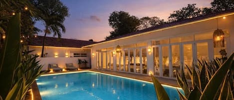 Uma piscina com cobertura de vidro, cercada por plantas e um edifício com grandes janelas.