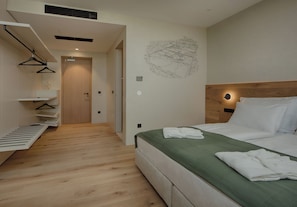 Quarto de hotel moderno com cama, piso de madeira, aquecedor fixado na parede e um cabideiro.
