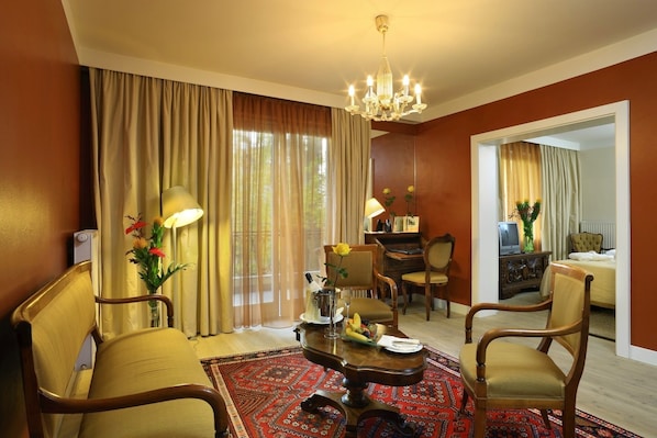 Quarto de hotel com área de jantar, um lustre, uma janela grande com cortinas e um espelho refletindo o ambiente.