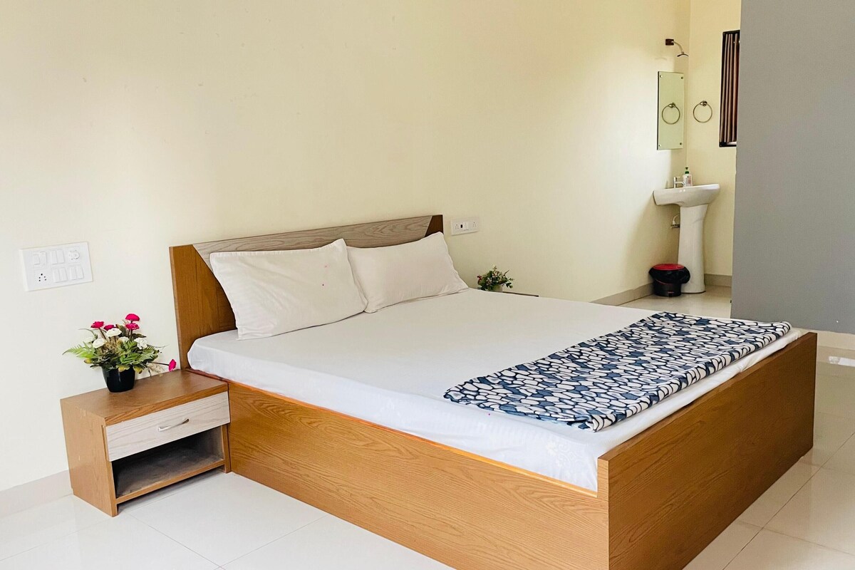 Quarto com cama de madeira, roupa de cama branca, mesa de cabeceira com planta, mesa auxiliar pequena e banheiro com pia e espelho.