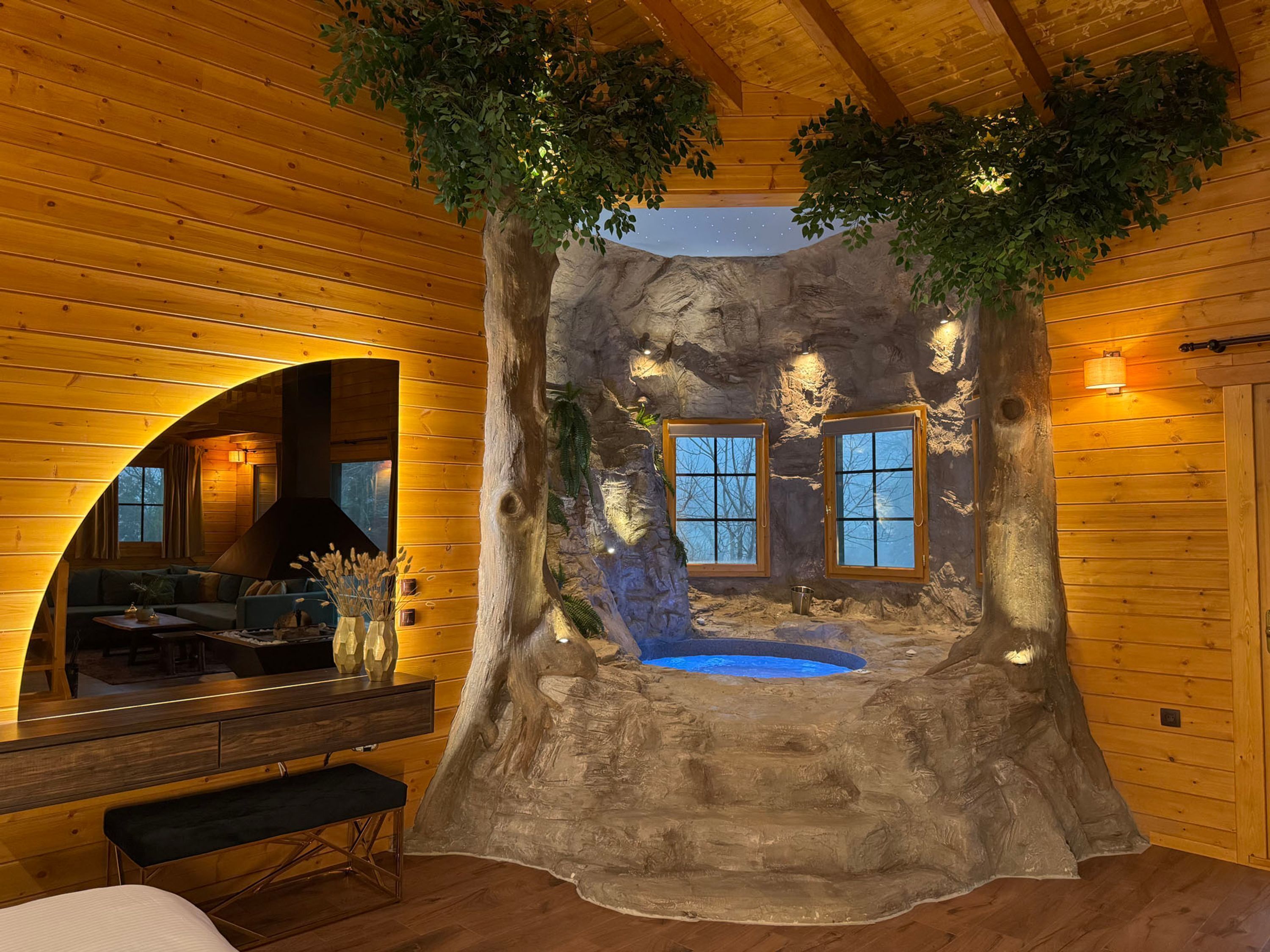 Interior de uma cabana de tronco com uma formação rochosa central, uma área de estar aconchegante e um pequeno lago.