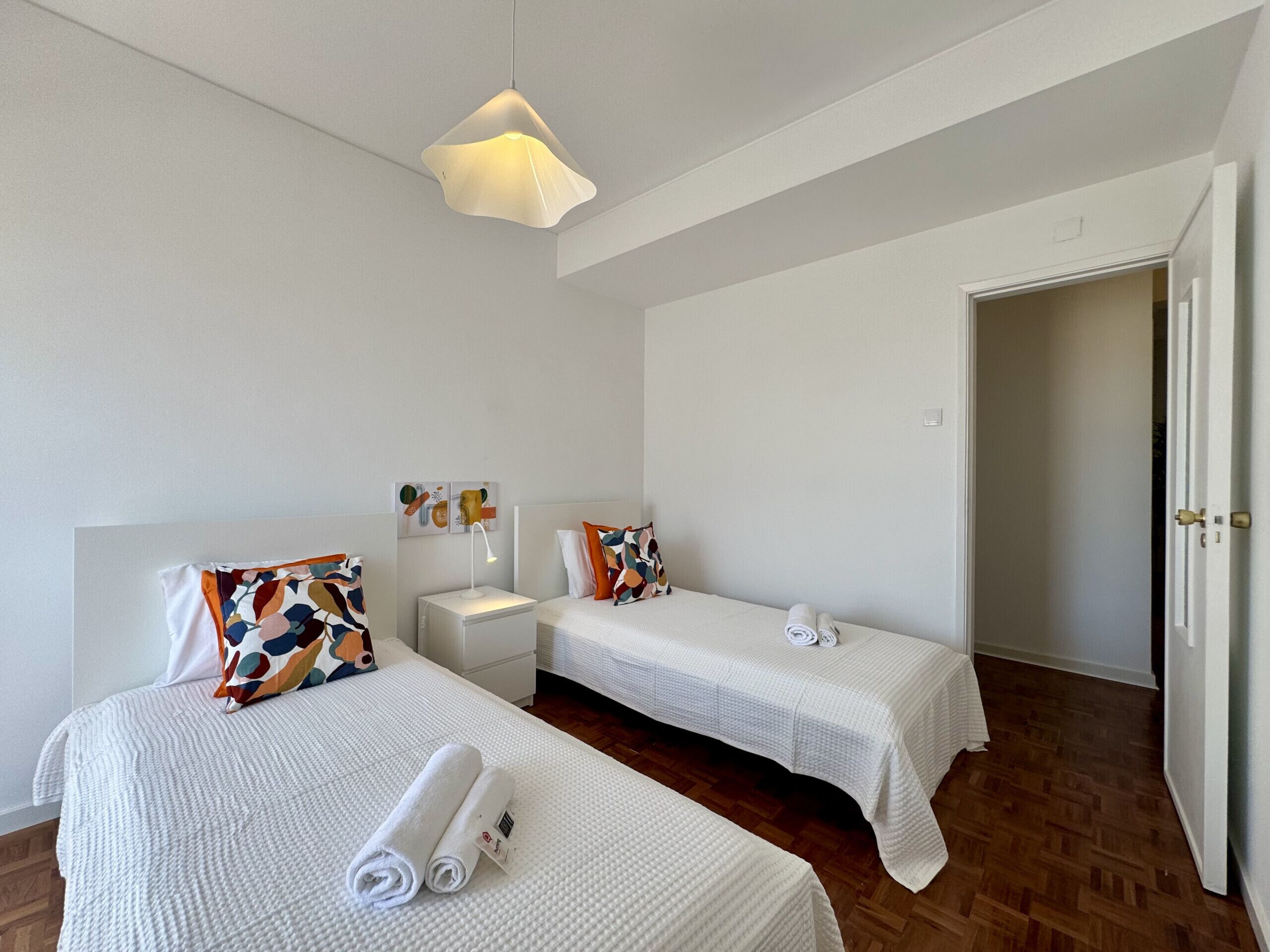 Quarto de hotel com duas camas, uma mesa de cabeceira, um abajur e uma porta.