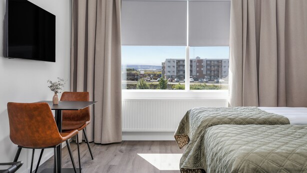 Quarto de hotel com cama, televisão na parede, mesa de jantar com cadeiras e vista para edifícios pela janela.