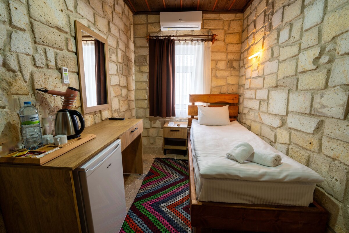 Quarto compacto com escrivaninha de madeira, cama, mesinha e janela com cortinas.