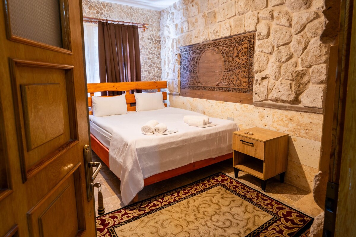 Quarto com parede de pedra, cama de madeira, mesa de cabeceira e tapete estampado.