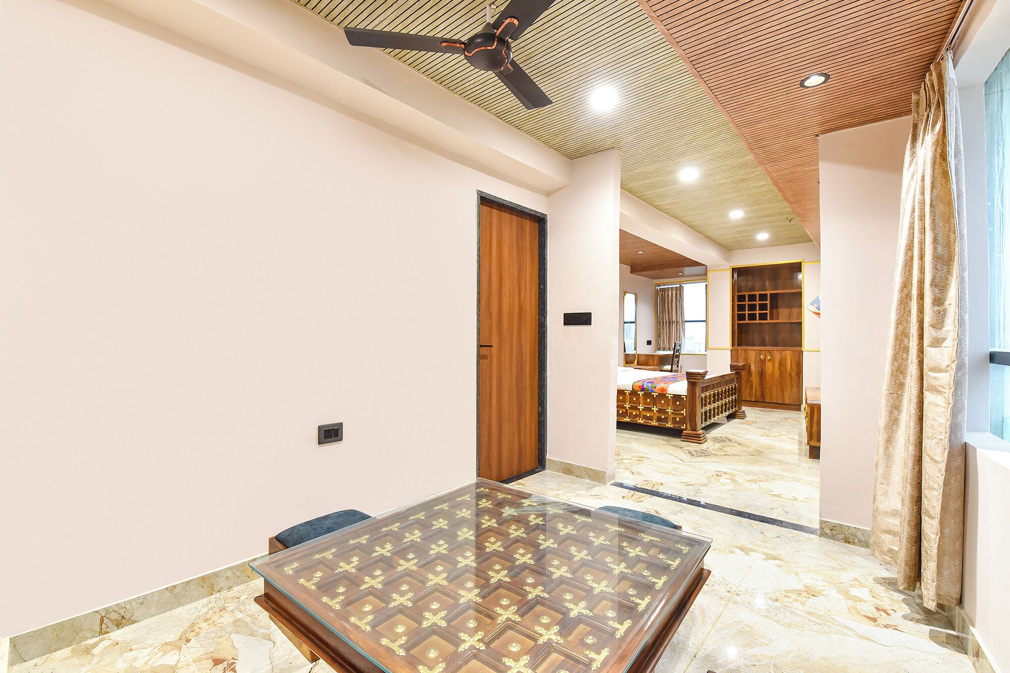 Quarto de hotel com mesa de vidro, ventoinha no teto, guarda-roupa de madeira e vista para o quarto vizinho.