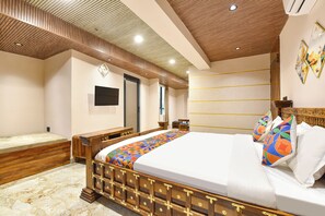 Quarto de hotel com uma cama grande, cabeceira de madeira, uma televisão fixada na parede e uma janela com cortinas.