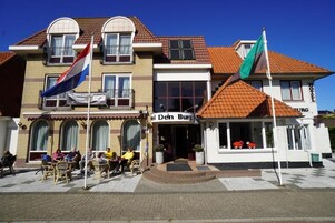Um hotel com uma placa indicando 'Hotel Den Burg', apresentando um estilo arquitetônico europeu tradicional, com telhado de telhas e varanda com guarda-corpo.