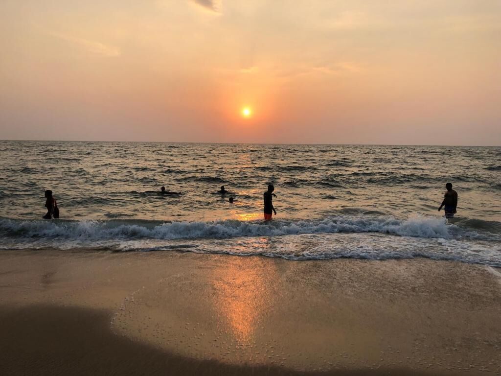 Cena de praia com o sol se pondo sobre o oceano, pessoas na água e uma faixa de areia.