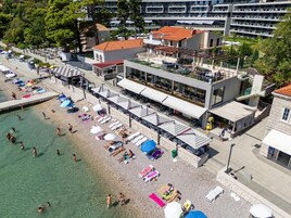Vista aérea de uma área costeira com uma praia, um restaurante com mesas ao ar livre e um edifício moderno com telhado vermelho.
