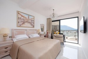 Quarto de hotel com uma cama grande, uma pintura de paisagem urbana, um candelabro, uma varanda com vista e uma TV de tela plana.