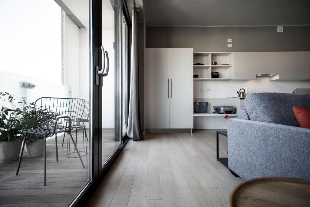 Uma sala de estar moderna com um sofá cinzento, uma área de jantar com mesa e cadeiras, e uma cozinha com armários e eletrodomésticos.