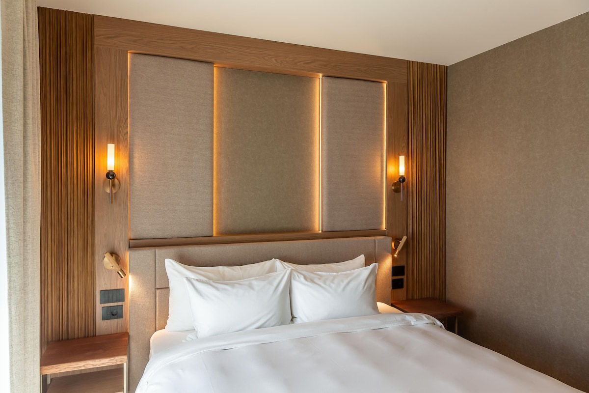 Quarto de hotel com cama grande, cabeceira com iluminação embutida e abajures laterais.