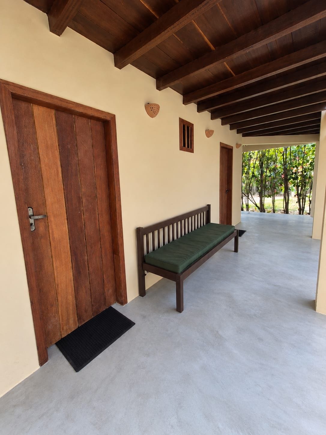 Área externa coberta com banco de madeira, uma porta e vista para o verde.