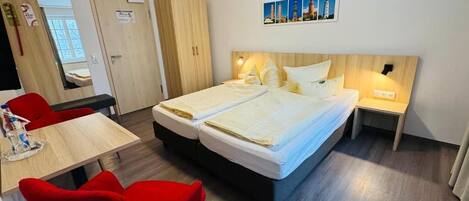 Quarto de hotel com uma cama, escrivaninha, cadeiras vermelhas, guarda-roupa e um quadro na parede.