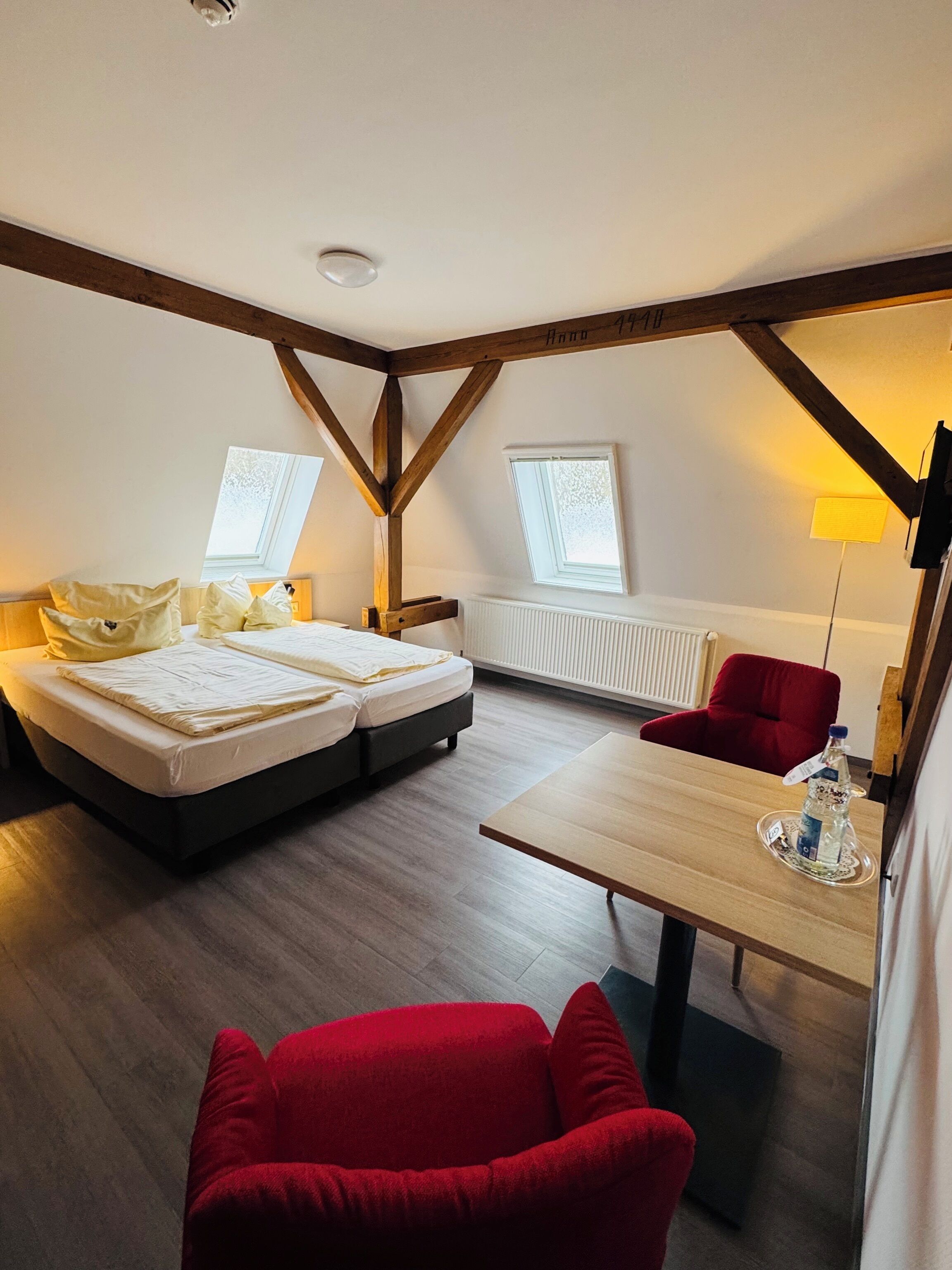 Quarto com uma cama, uma cadeira vermelha, uma mesa com uma garrafa e duas janelas.