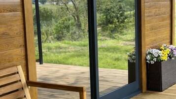 Um deck de madeira com um banco, uma porta de vidro que dá para vista de árvores e gramado, e um vaso com flores.