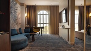 Quarto de hotel com sofá curvo, uma pequena mesa, uma cadeira, televisão fixada na parede e uma janela grande com cortinas.