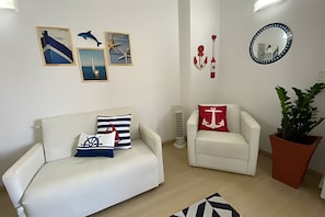 Uma sala de estar com um sofá branco, uma poltrona menor e uma planta em vaso. Há decorações nas paredes, incluindo quadros e um espelho.