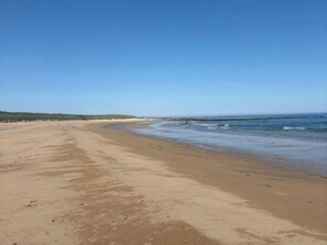 Uma extensa praia arenosa com ondas suaves, céu claro e um afloramento rochoso ao longe.