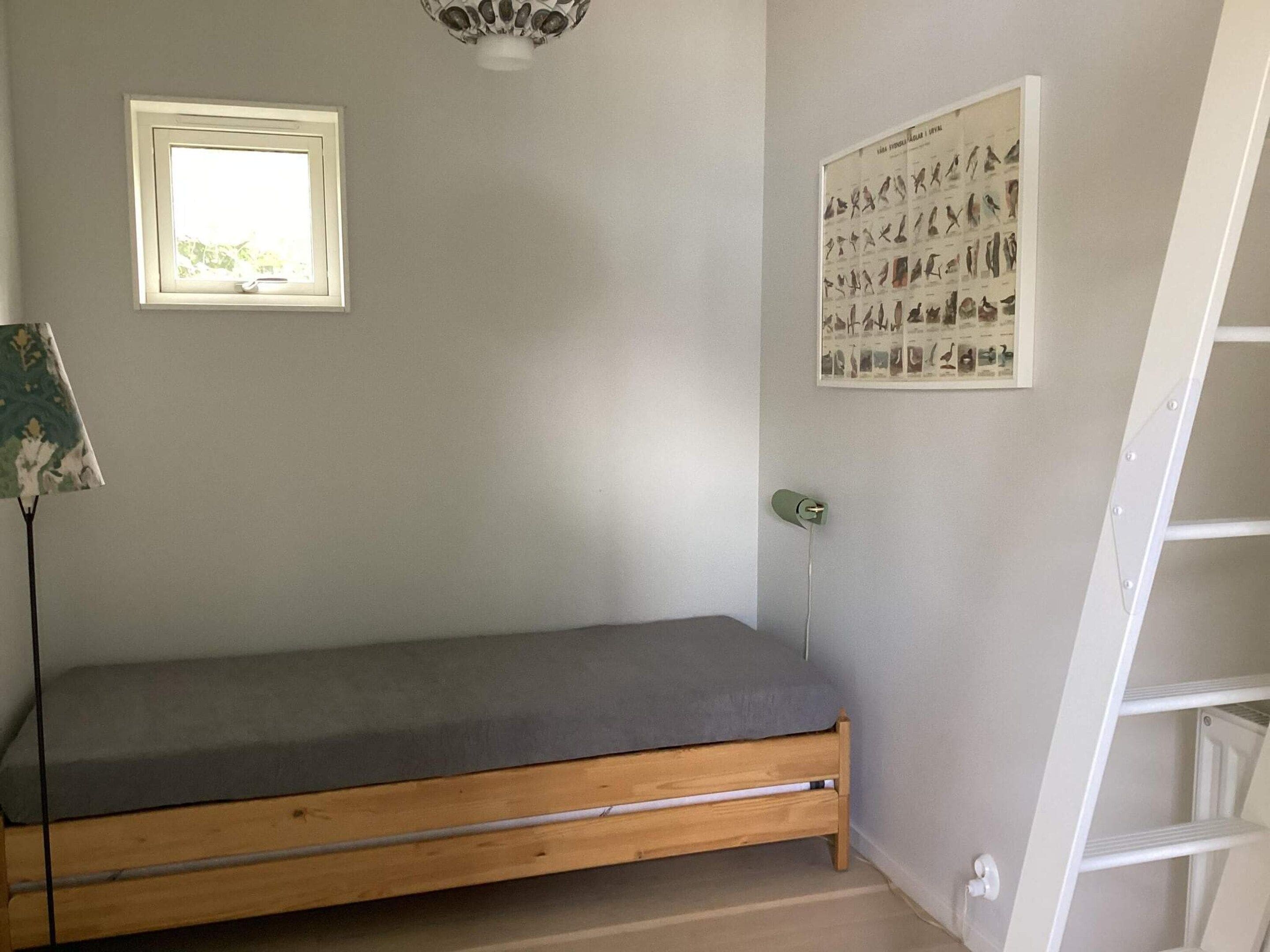 Quarto pequeno com cama de solteiro, estrutura de madeira, janela, luminária na parede e ilustração de um pássaro.