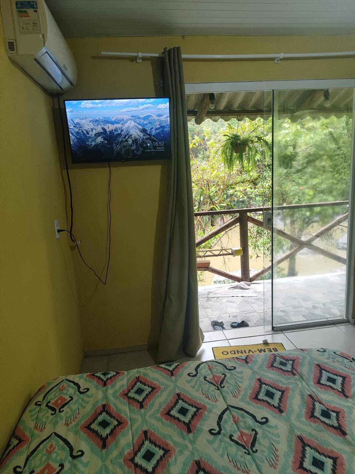 Quarto com uma cama, televisão presa na parede e porta de vidro deslizante para uma área externa.