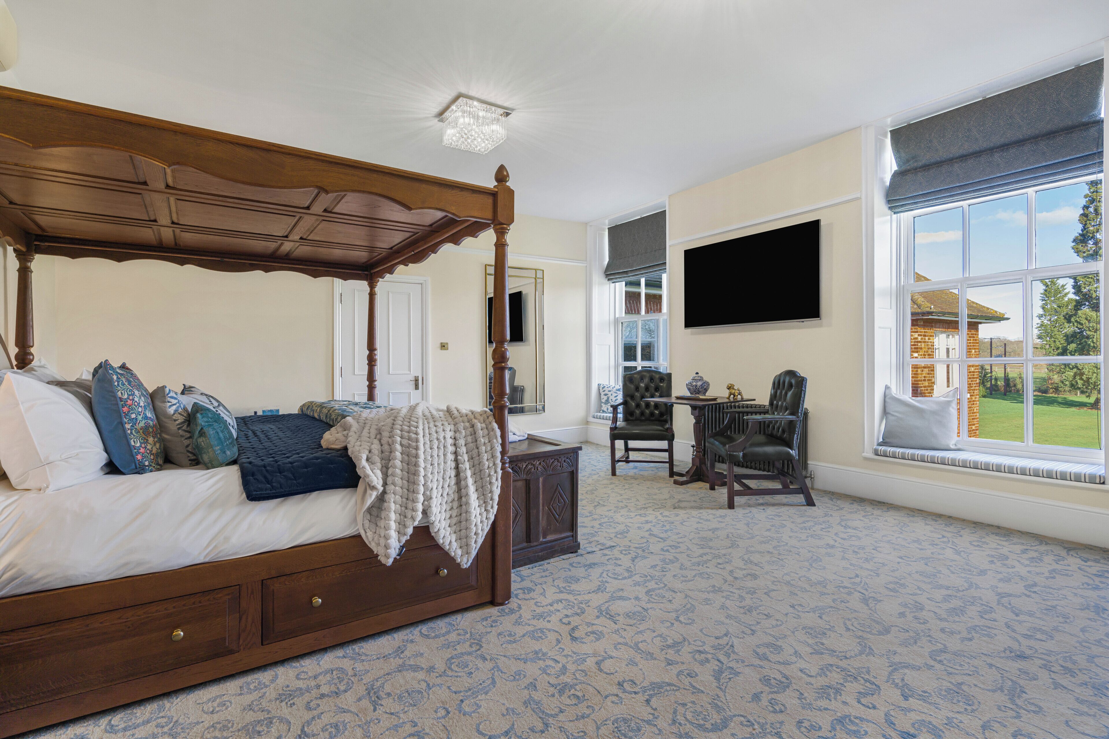 Uma cama de dossel com cabeceira de madeira, uma mesinha de cabeceira e uma poltrona com assento acolchoado.