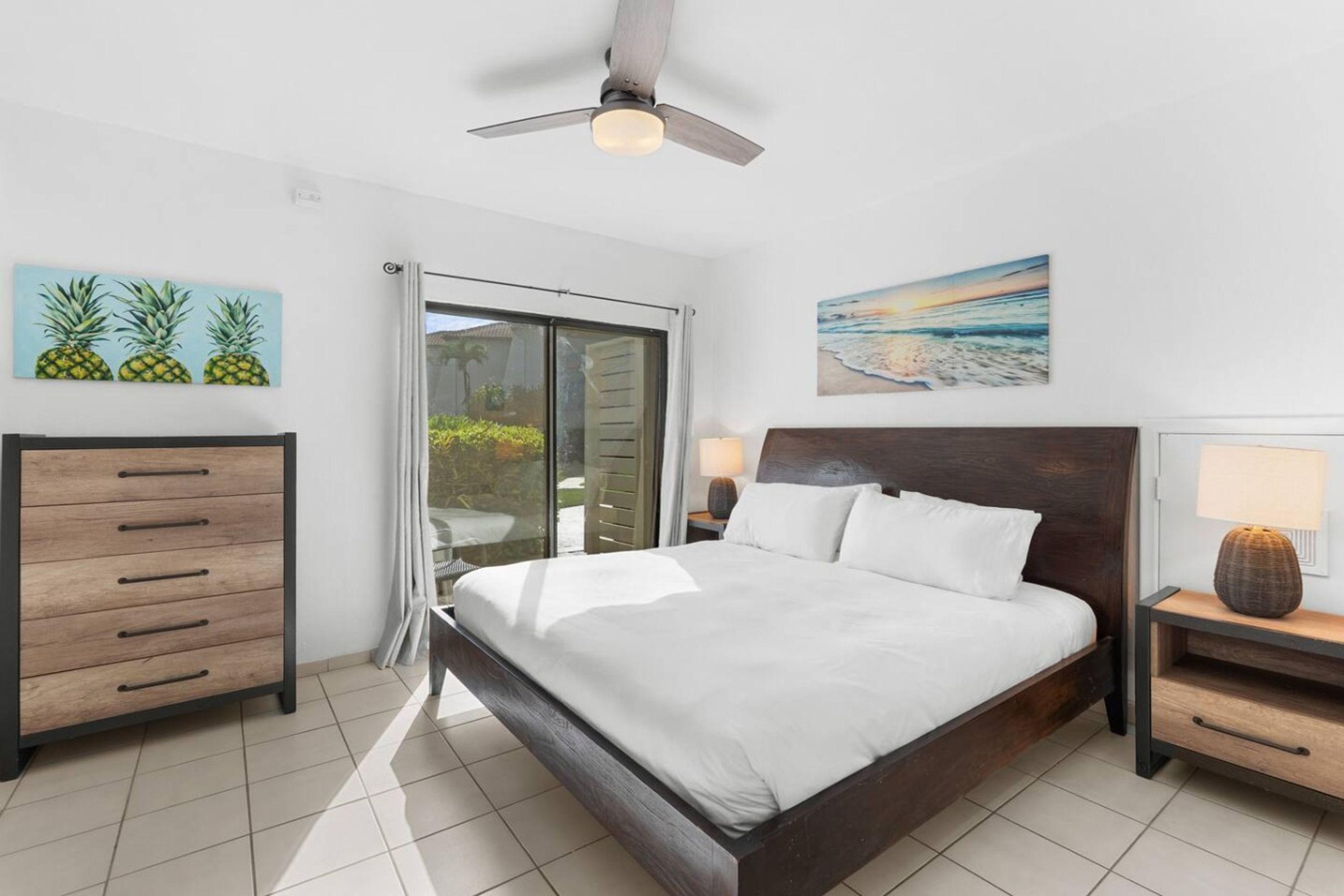 Quarto com cama, mesa de cabeceira, cômoda, ventilador de teto e porta de vidro corrediça que dá para área externa.