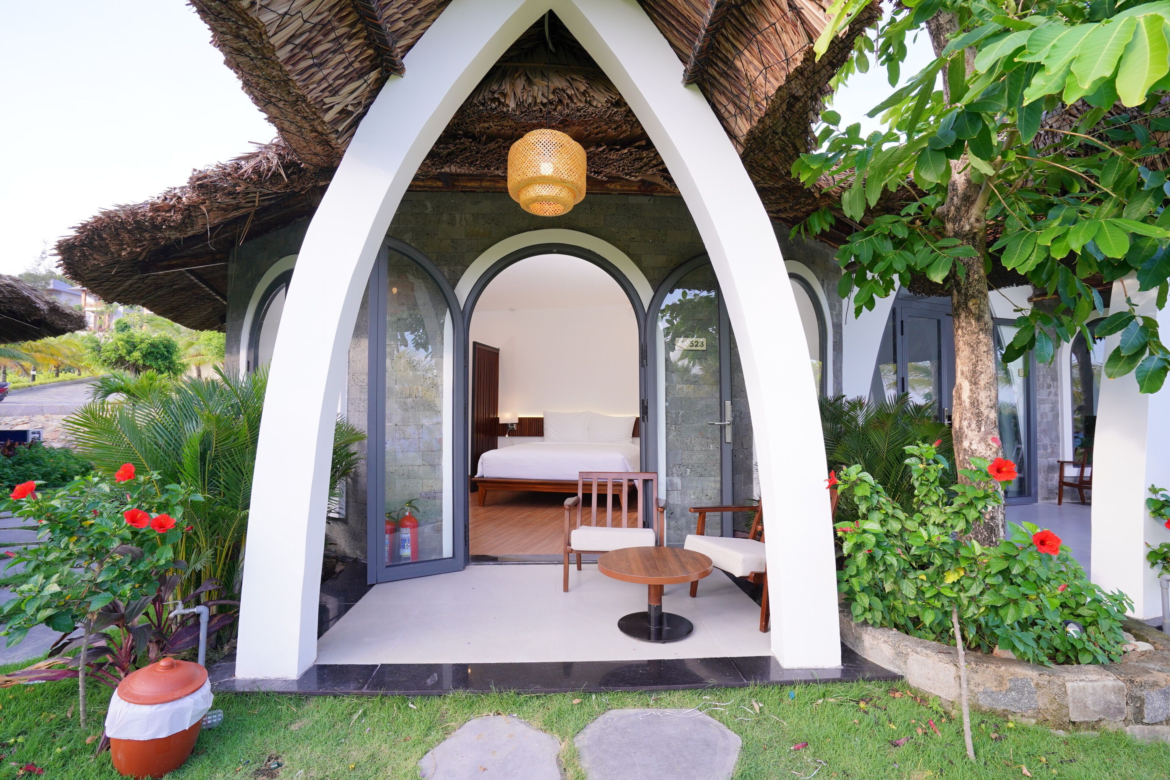 Uma villa moderna com telhado de colmo, um quarto com cama e área de estar, e um jardim com vasos de plantas e flores.