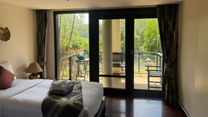 Quarto de hotel com cama, mesa de cabeceira, luminária e vista para um pátio externo com mesa e cadeiras.