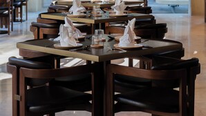 Restaurante com mesas postas para refeições, vista para o verde através de grandes janelas e luminárias pendentes.