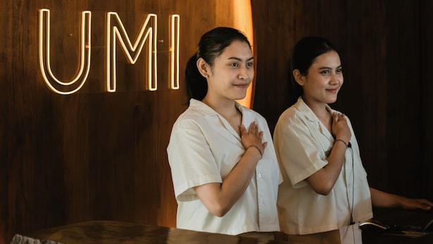 Duas pessoas em um balcão de recepção, com um fundo de madeira e letras iluminadas que formam a palavra 'UMI'.