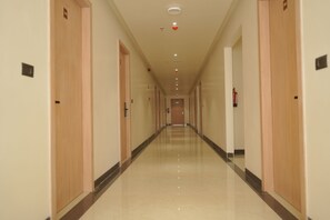 Corredor longo e estreito de hotel com portas de madeira em ambos os lados, piso revestido de azulejos brilhantes e teto com iluminação embutida.