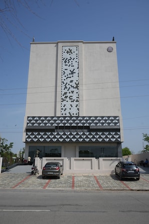 Um edifício com um grande painel vertical de padrão geométrico, telhado plano e área de estacionamento com dois carros.