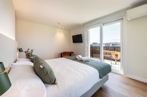 Quarto de hotel com cama, televisão, varanda com vista para edifícios e chão de madeira.