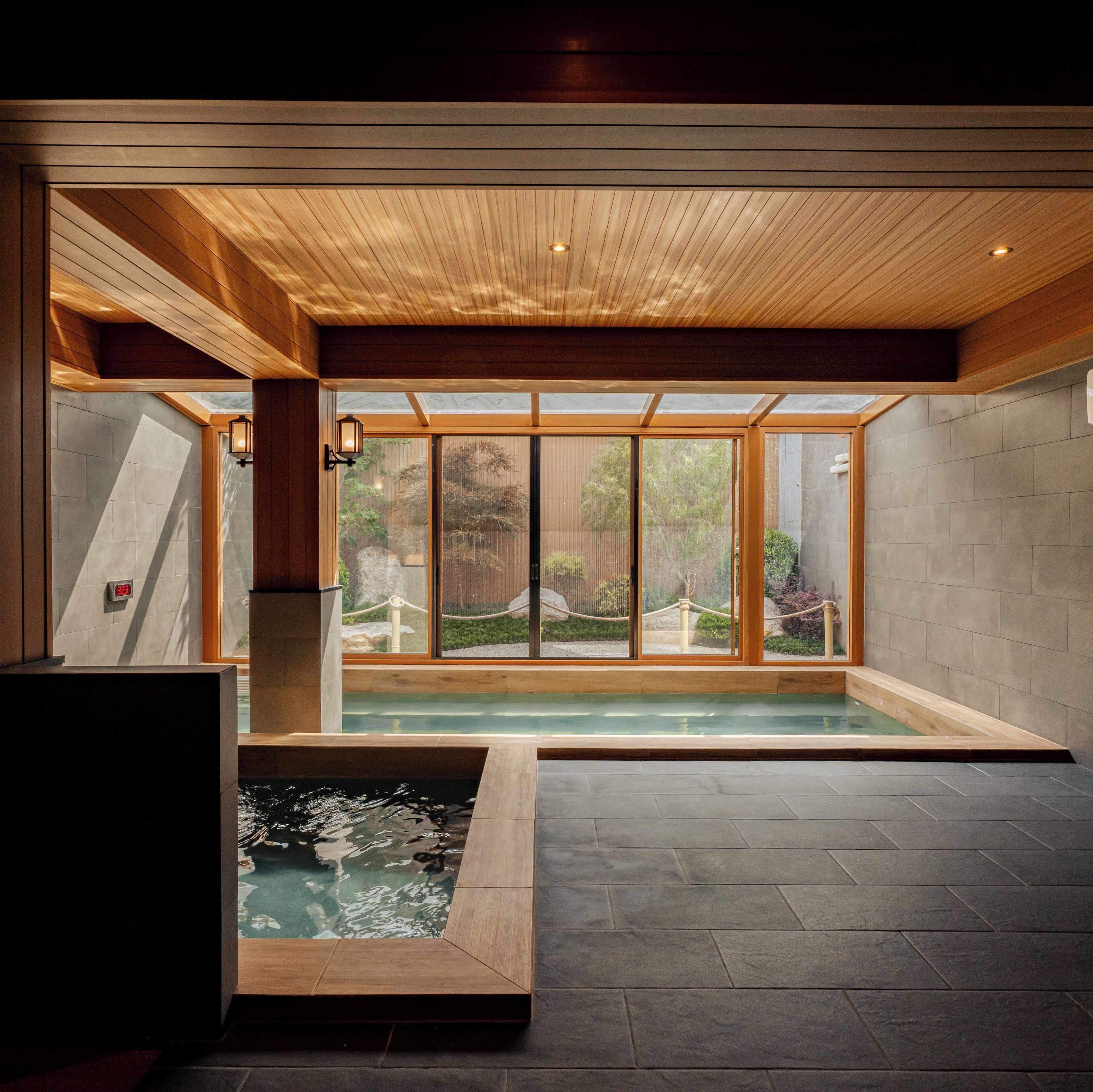 Uma área interna de piscina moderna, com teto de madeira, paredes de pedra e vista para um jardim.