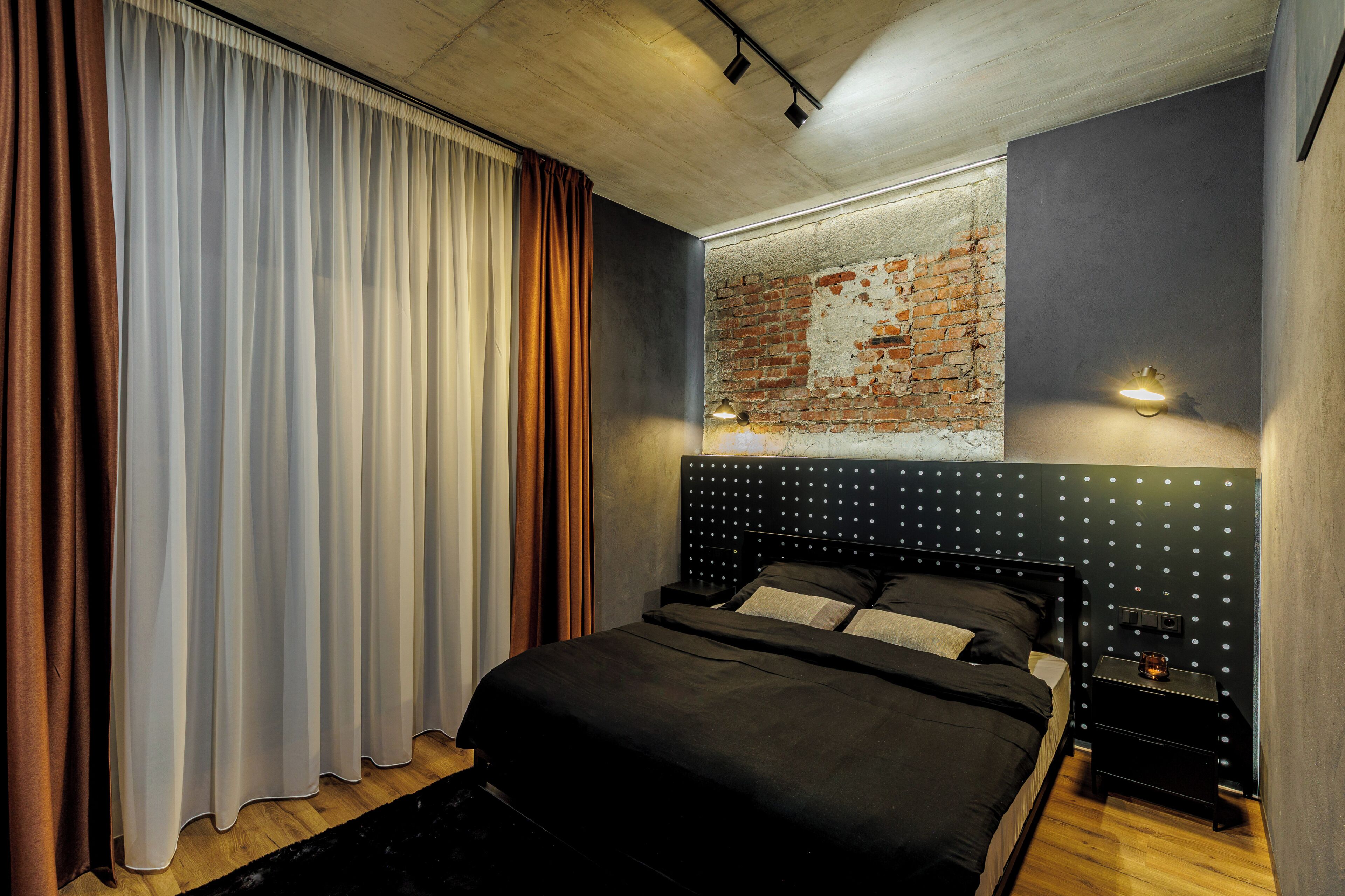 Quarto com cama, cortinas, parede de tijolos e mesa de cabeceira com abajur.