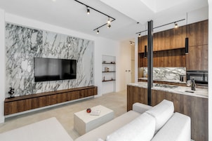Uma sala de estar moderna com uma parede de mármore, uma TV de tela plana, um console de madeira, um sofá branco e uma área de cozinha com armários de madeira e bancada de mármore.