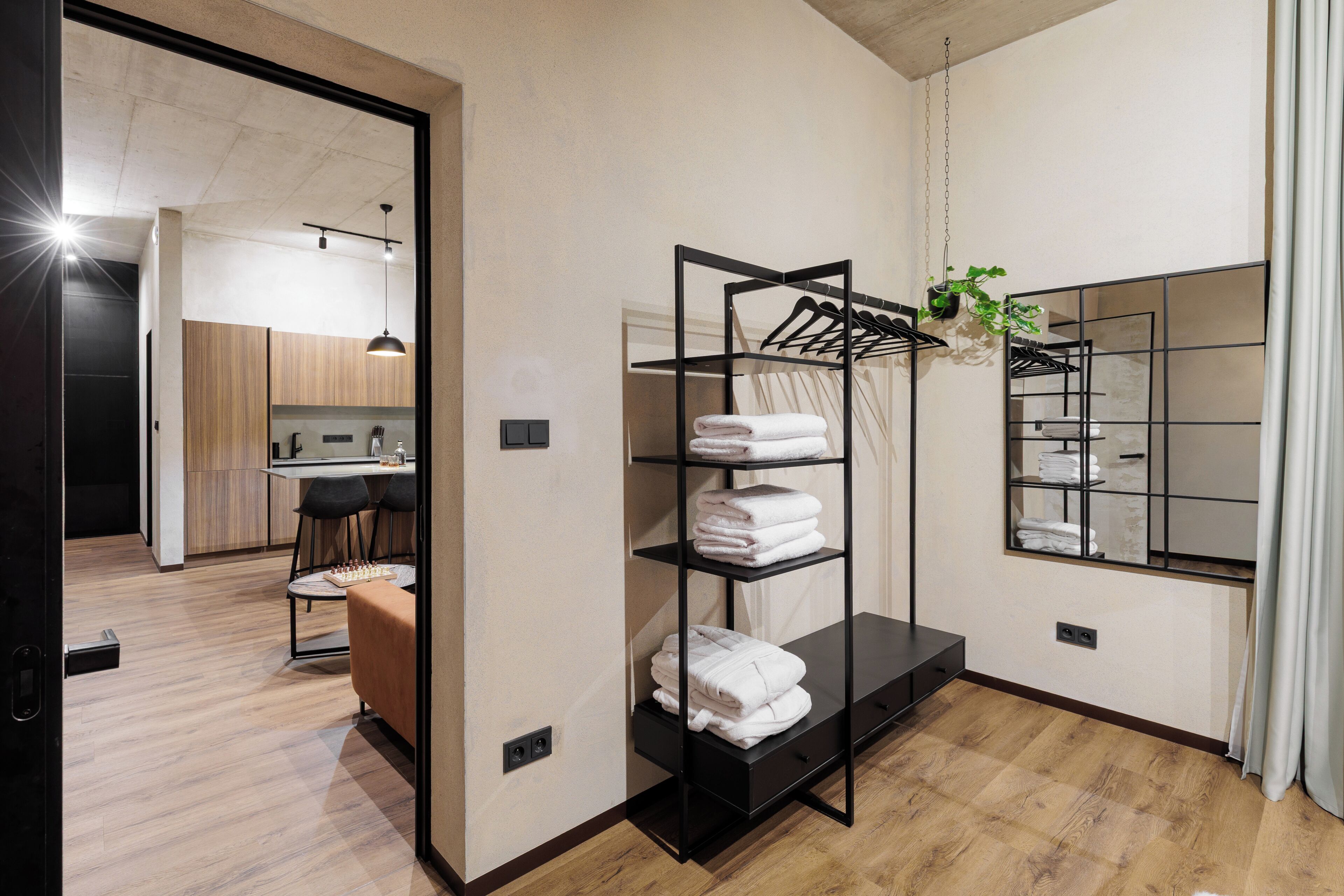 Quarto moderno com piso de madeira, uma estante preta com toalhas, uma planta pendente e uma janela com cortinas.
